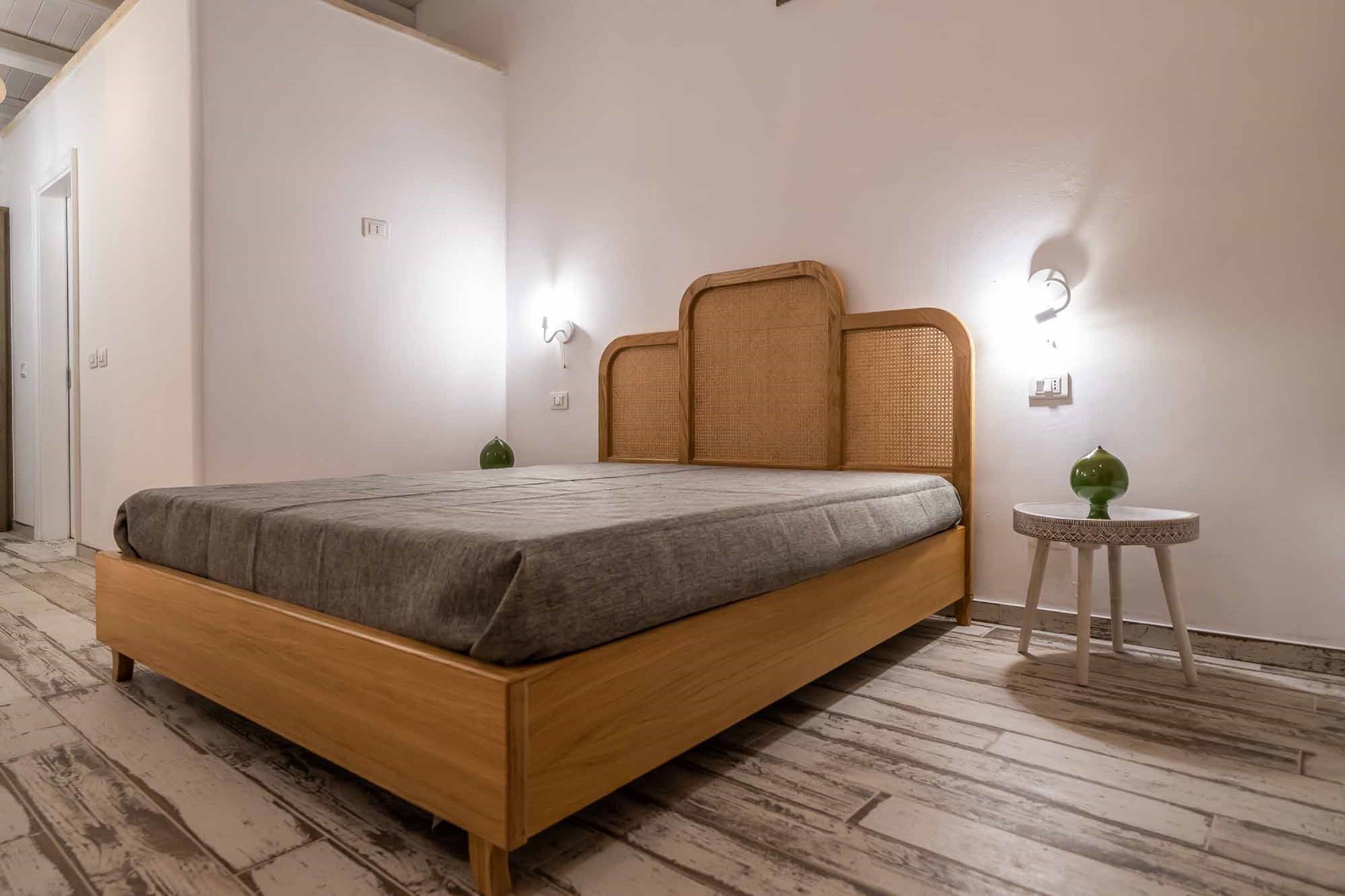 Quarto moderno com cama de madeira, duas luminárias na parede e uma pequena mesa redonda com um vaso verde.