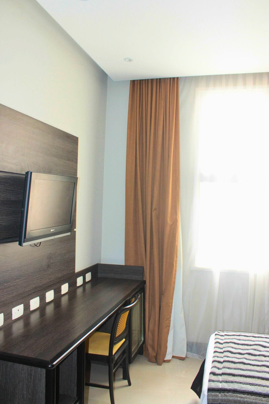 Quarto de hotel com escrivaninha de madeira escura, televisão na parede, uma cadeira, cama com manta listrada e uma janela grande com cortinas.