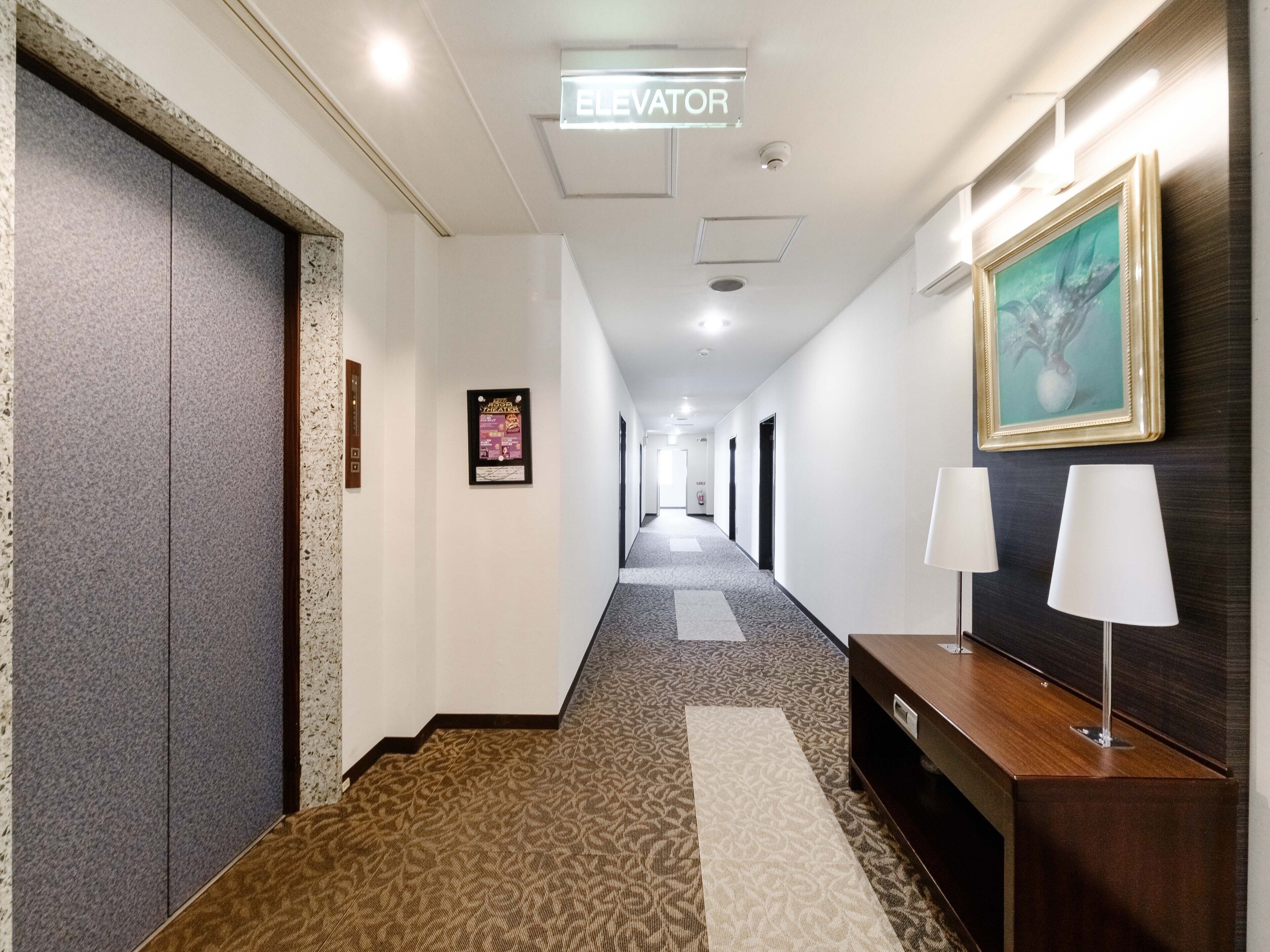 Um corredor de hotel com piso de carpete, uma escrivaninha de madeira com luminárias, um quadro pendurado na parede e o sinal de elevador.