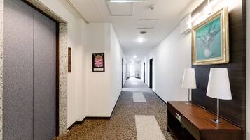 Um corredor de hotel com piso de carpete, uma escrivaninha de madeira com luminárias, um quadro pendurado na parede e o sinal de elevador.
