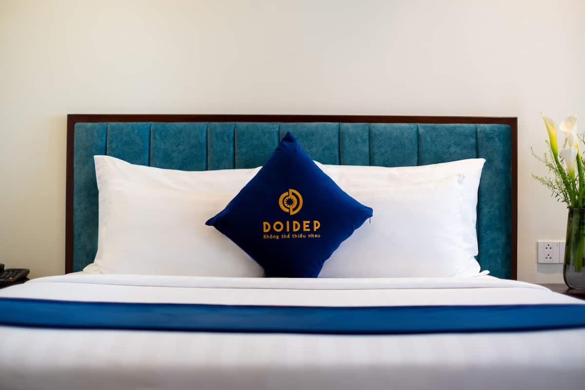Cama bem arrumada com cabeceira azul, roupa de cama branca e travesseiro azul com logotipo.