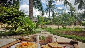 Mesa de madeira com duas refeições e duas canecas em varanda, com vista para um jardim e palmeiras.