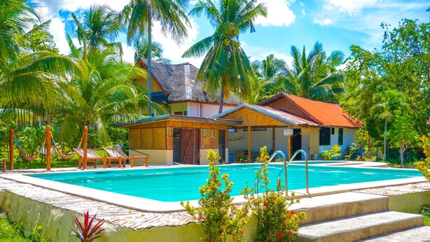 Uma área de piscina com piscina azul cristalina, cercada por palmeiras e um edifício com telhado vermelho.