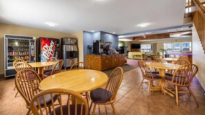 Restaurante com mesas e cadeiras de madeira, área de cozinha e máquina de vendas automática.
