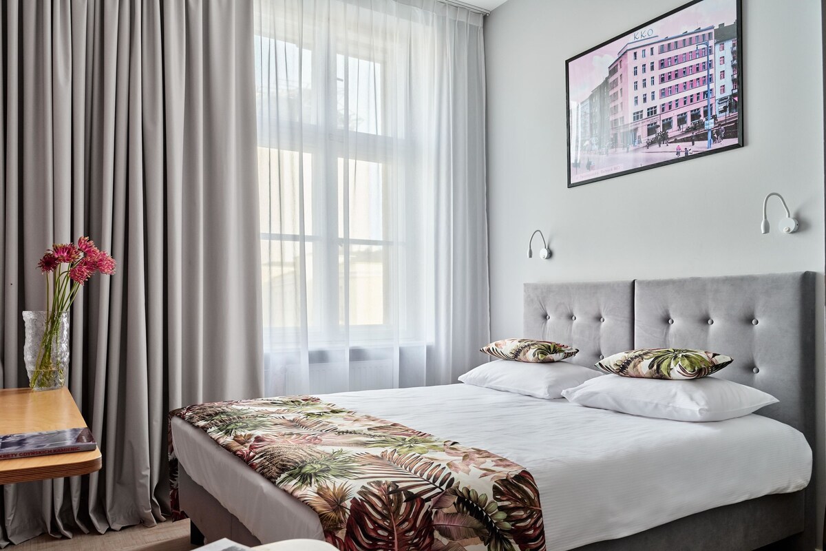 Quarto de hotel com uma cama grande, uma mesa de cabeceira com um livro, um vaso com flores e um quadro na parede.
