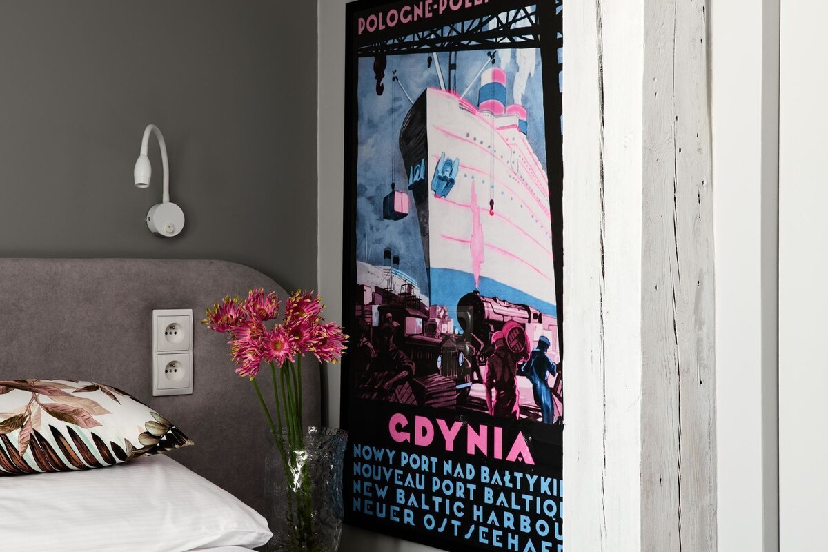 Um quarto com uma cama, uma mesa de cabeceira, uma luminária na parede, um pôster e um vaso com flores.