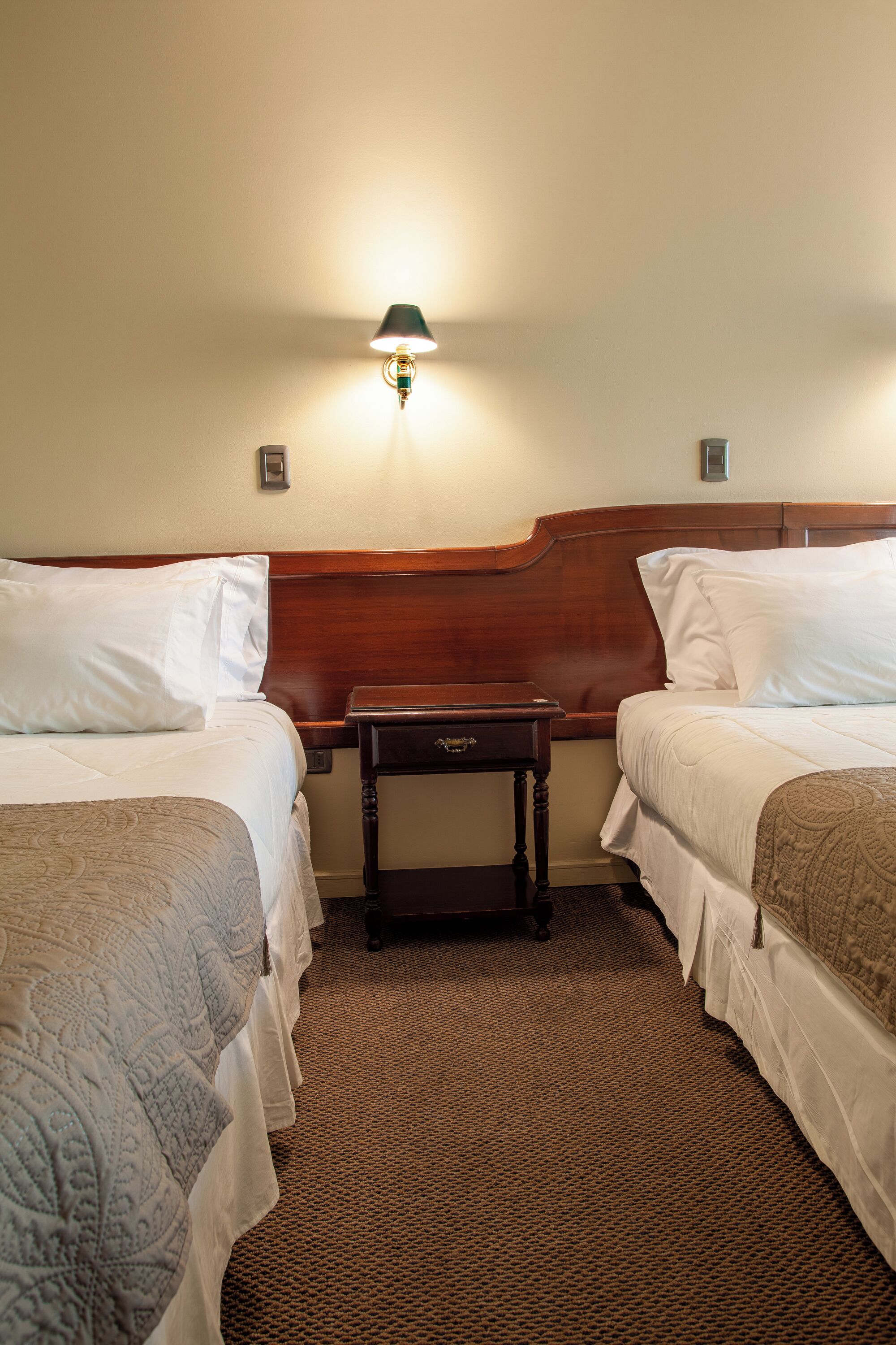 Quarto de hotel com duas camas, uma cabeceira de madeira, mesa de cabeceira e uma luminária fixada na parede.