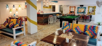 Uma sala de bilhar com mesa de bilhar, espaço de bar e assentos com almofadas coloridas em zigue-zague.