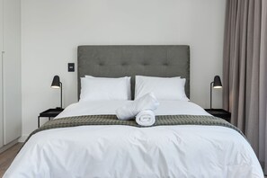 Cama bem arrumada com lençóis brancos, cabeceira cinza e duas luminárias laterais.