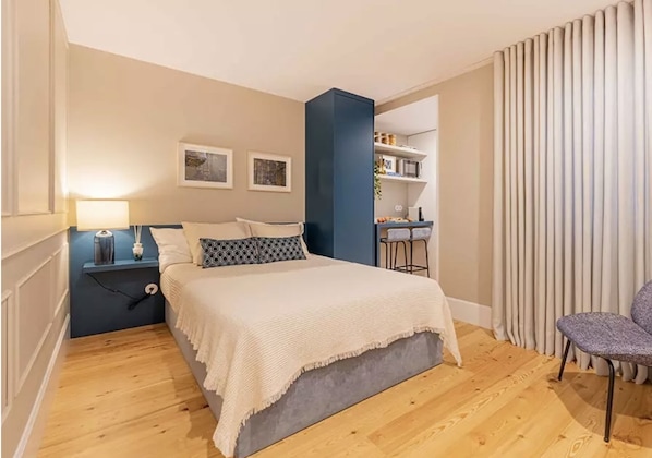 Quarto com cama, mesa de cabeceira, luminária e uma pequena área de cozinha.