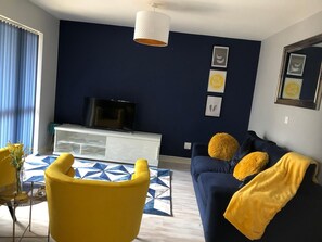 Uma sala de estar com parede azul escura, um móvel de entretenimento branco, uma poltrona amarela, um sofá azul com almofadas amarelas e um tapete estampado.