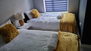 Quarto com duas camas, cada uma com um edredom branco e um travesseiro fofo. Há uma mesa de cabeceira com uma luminária e uma gaveta pequena.
