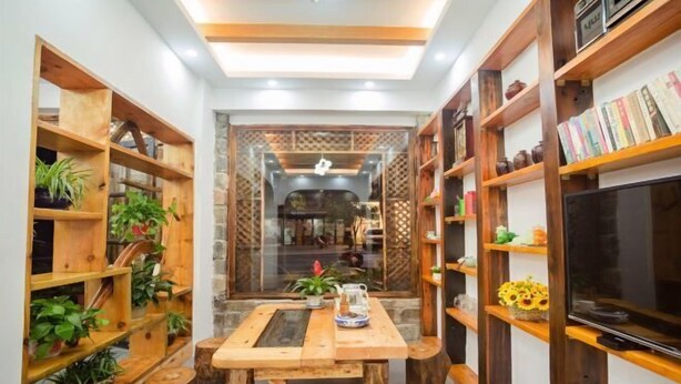 Um quarto aconchegante com estantes de madeira repletas de livros e plantas, uma mesa de madeira com uma chaleira, e uma TV à direita.