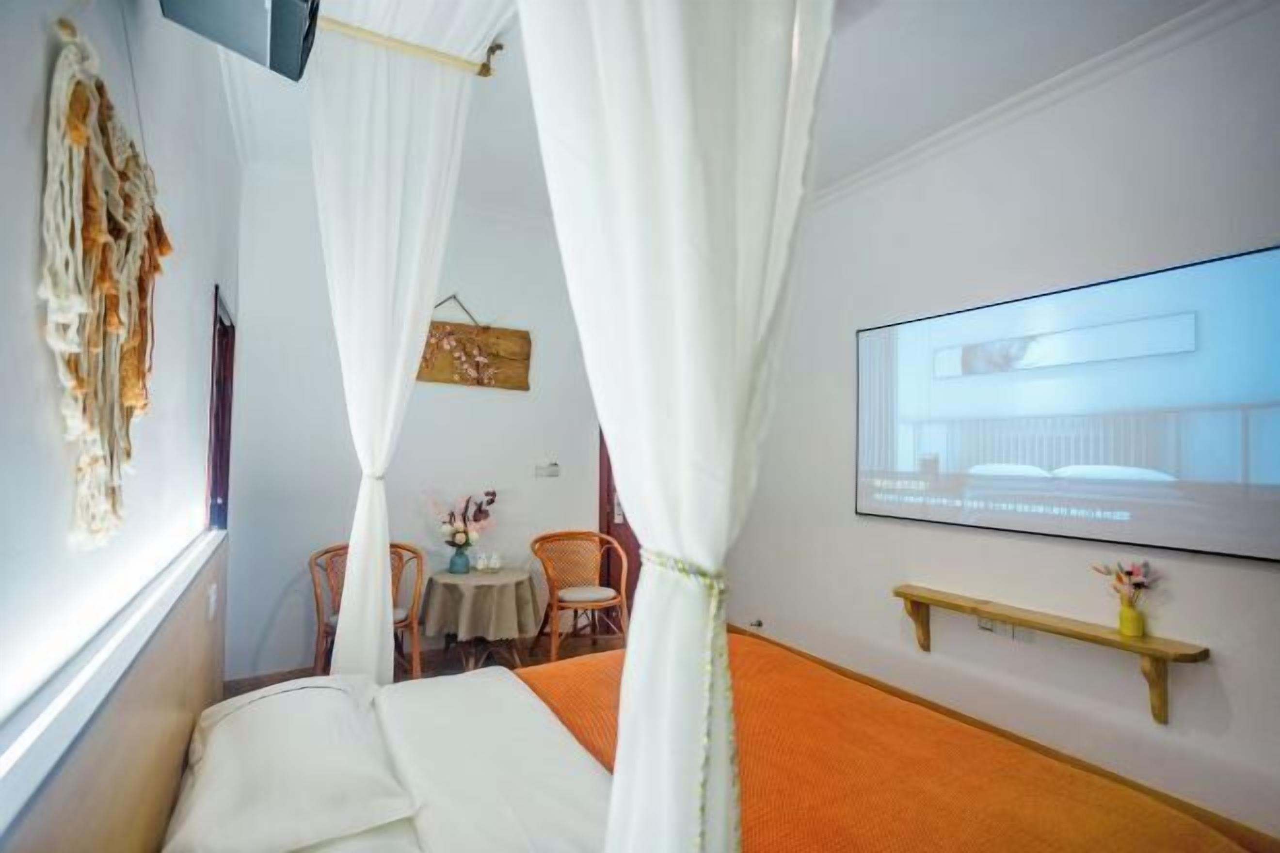 Quarto com cama, televisão presa na parede, mesinha com cadeiras e janela com cortinas.