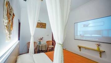 Quarto com cama, televisão presa na parede, mesinha com cadeiras e janela com cortinas.