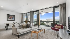 Uma sala de estar moderna com um sofá, uma mesa de centro e uma cadeira vermelha. Há uma janela grande com vista para prédios e céu.