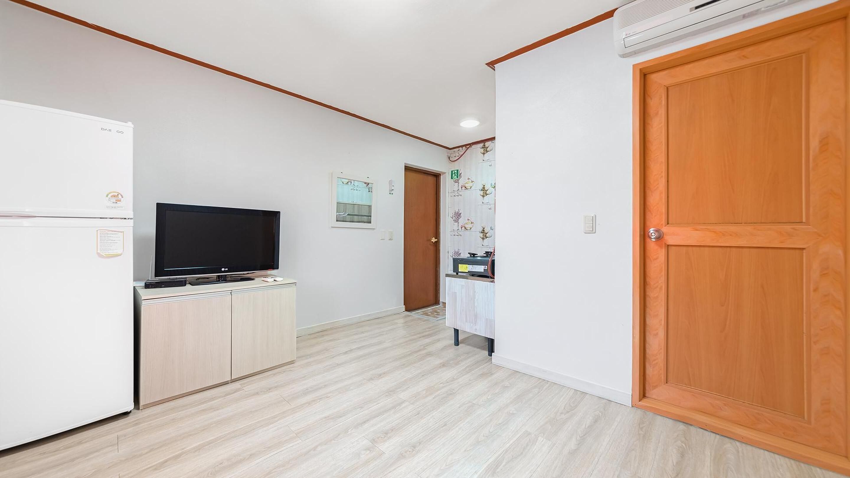 Quarto pequeno e limpo, com frigorífico, televisão sobre um móvel de madeira e porta de madeira.