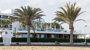 Um hotel à beira-mar com arquitetura moderna, palmeiras e uma praia de areia.