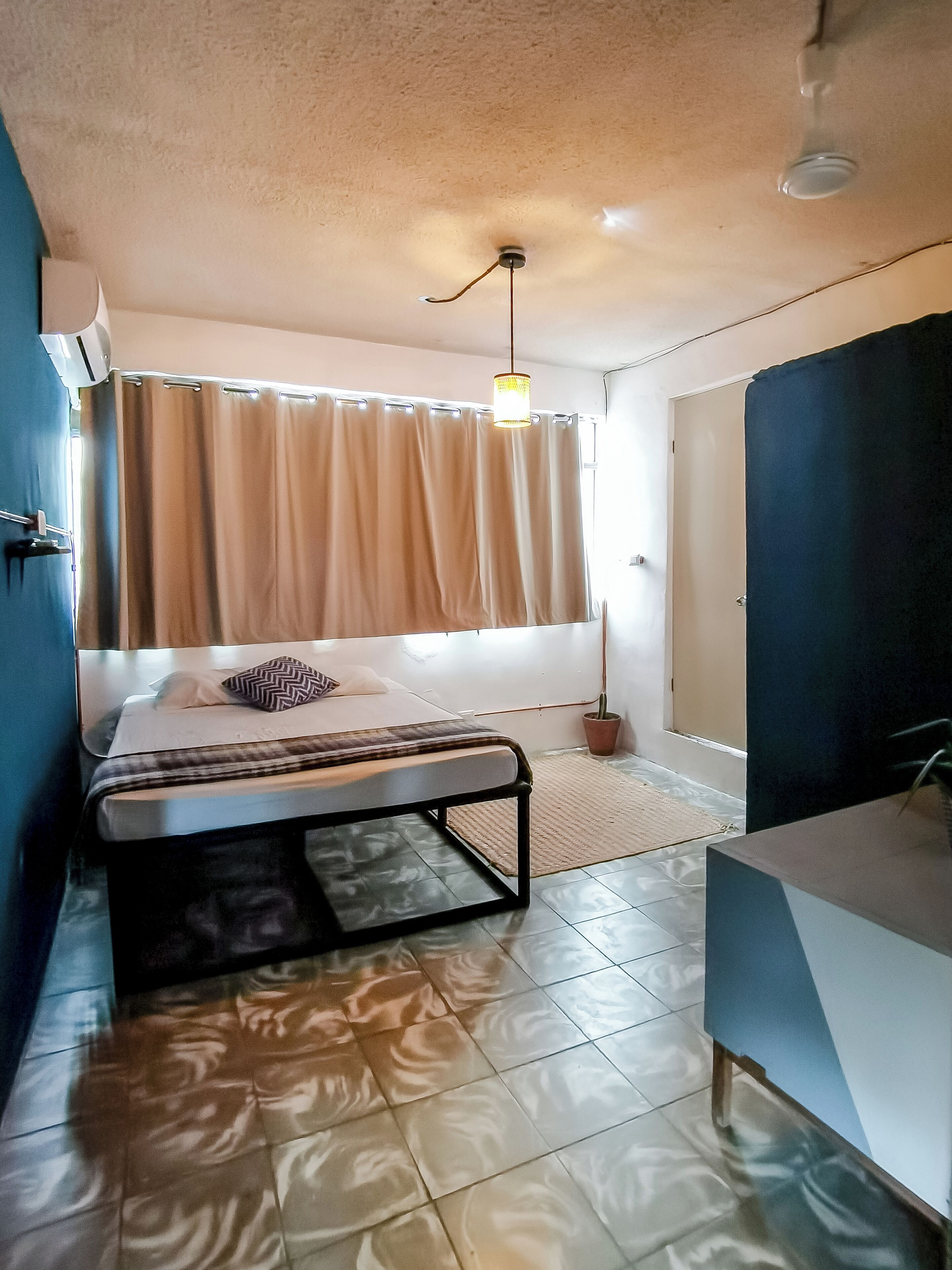 Quarto com cama, mesa de cabeceira, luminária pendente, janela com cortinas e um vaso de planta.