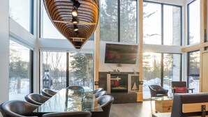 Um interior moderno com uma grande mesa de jantar, cadeiras de couro, lareira e amplas janelas com vista para o exterior.