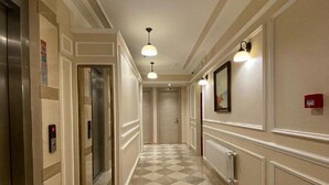 Corredor de hotel com piso xadrez, parede espelhada e porta com botão de elevador.