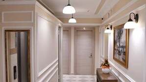 Um corredor com piso quadriculado, um banco de madeira, luminárias na parede e uma porta no final.
