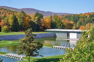 Um edifício moderno com telhado plano e janelas amplas, cercado por um lago e árvores com folhagem outonal.