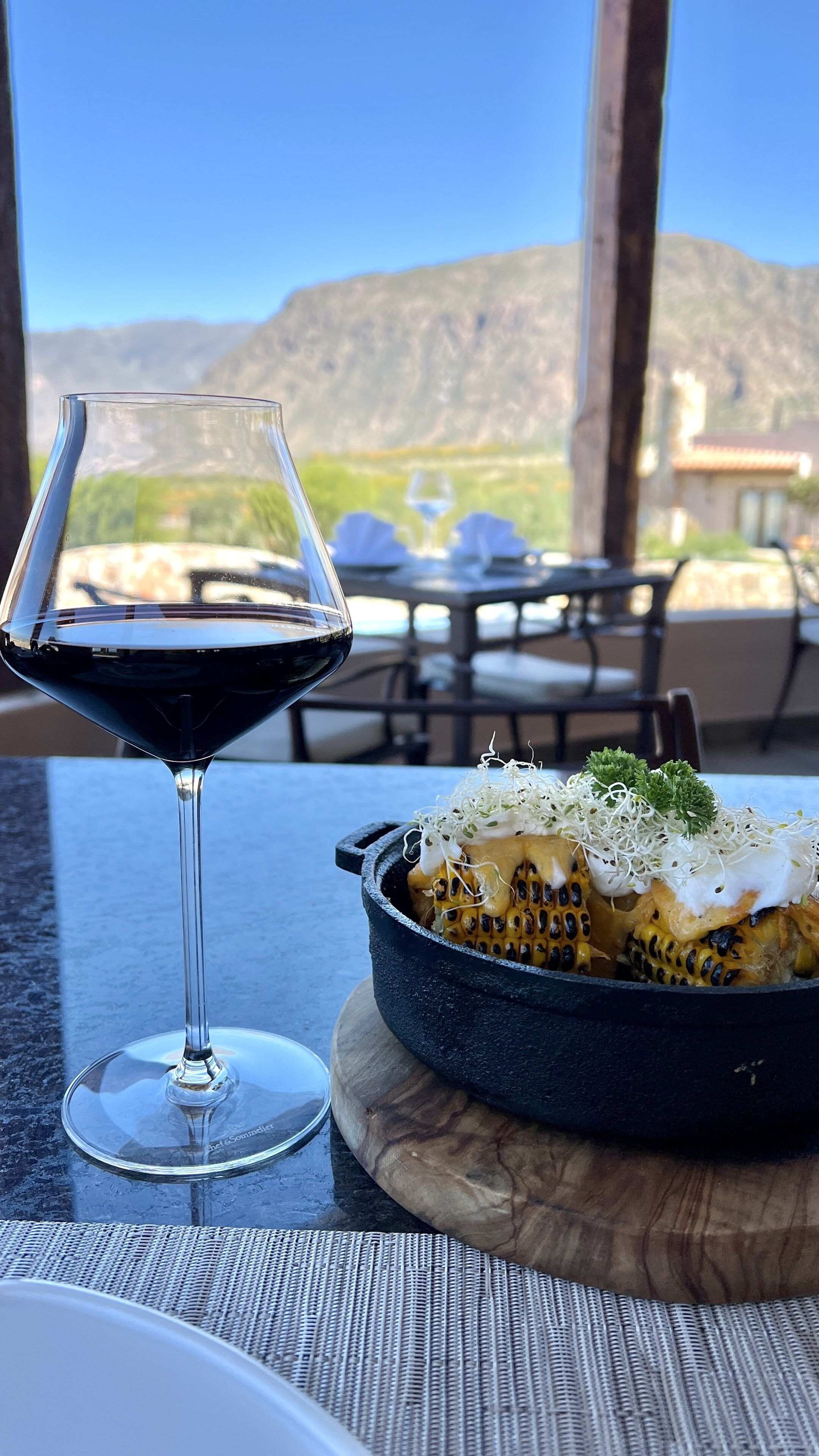 Um copo de vinho tinto sobre uma mesa, com um prato de milho grelhado coberto de brotos e ervas.