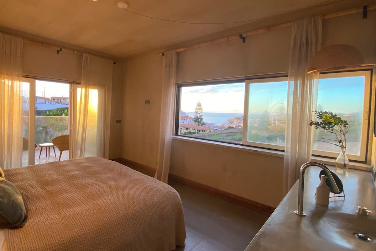 Quarto de hotel com cama, escrivaninha com pia, uma cadeira e grandes janelas com vista para a cidade.