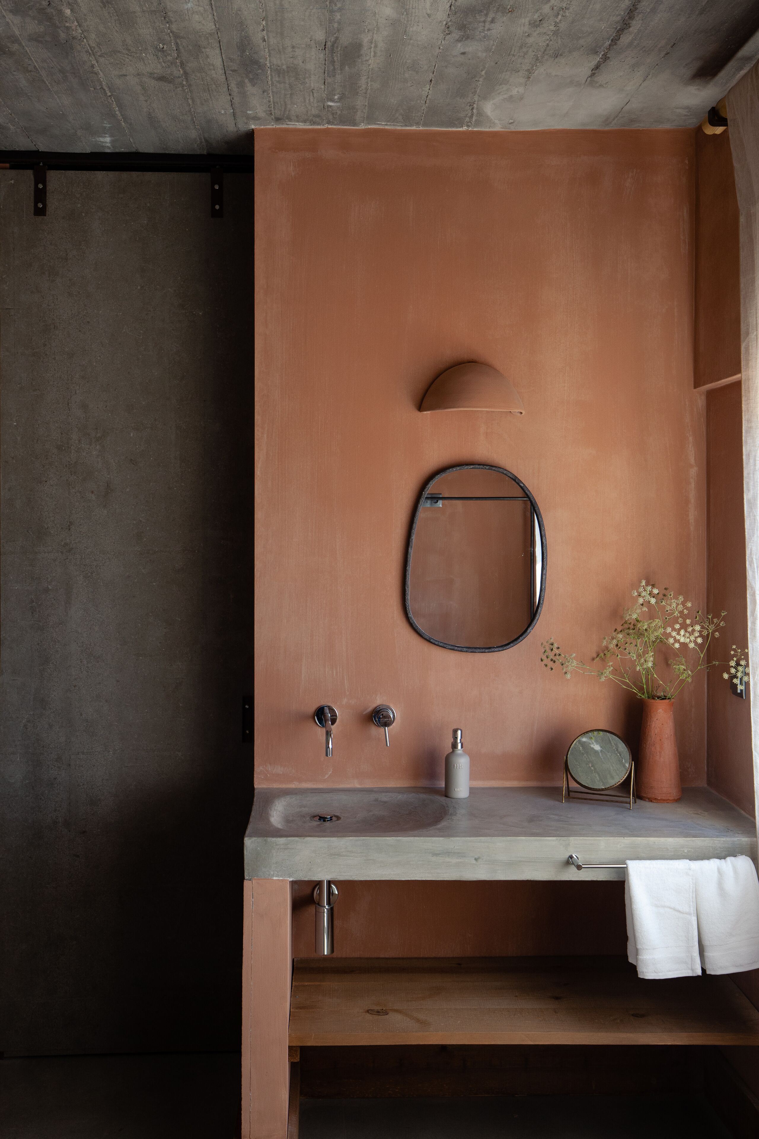 Casa de banho com teto de concreto, toucador de madeira, espelho redondo, lavatório e uma pequena planta em vaso.