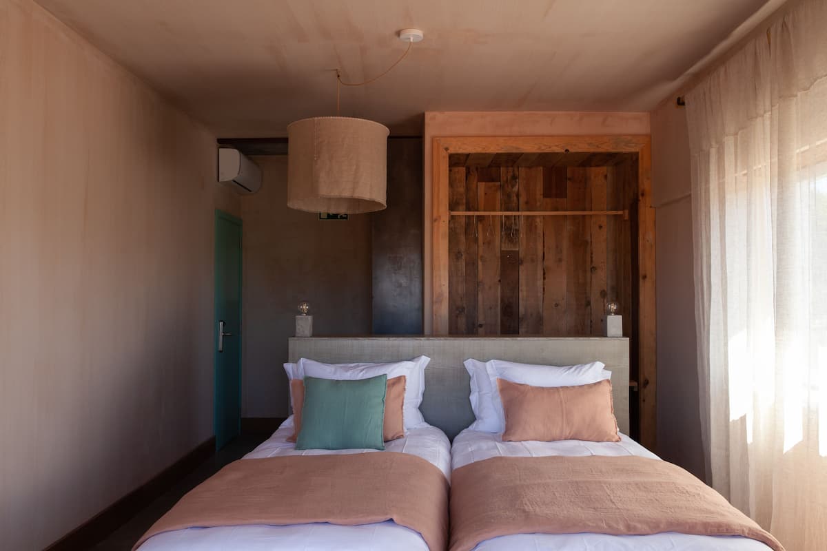 Quarto duplo com duas camas, cabeceira de madeira, luminária pendente e porta que dá para outro quarto.