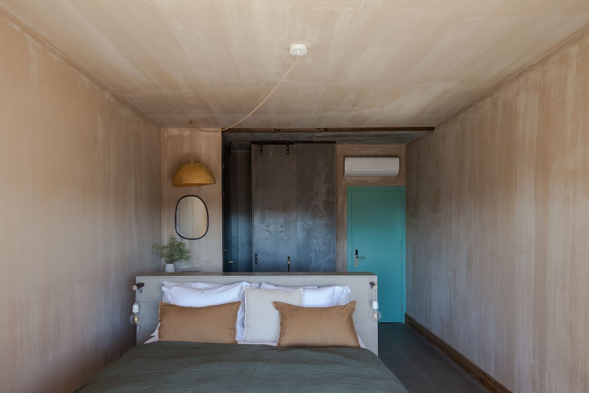 Quarto com cama, mesa de cabeceira, abajur e porta.