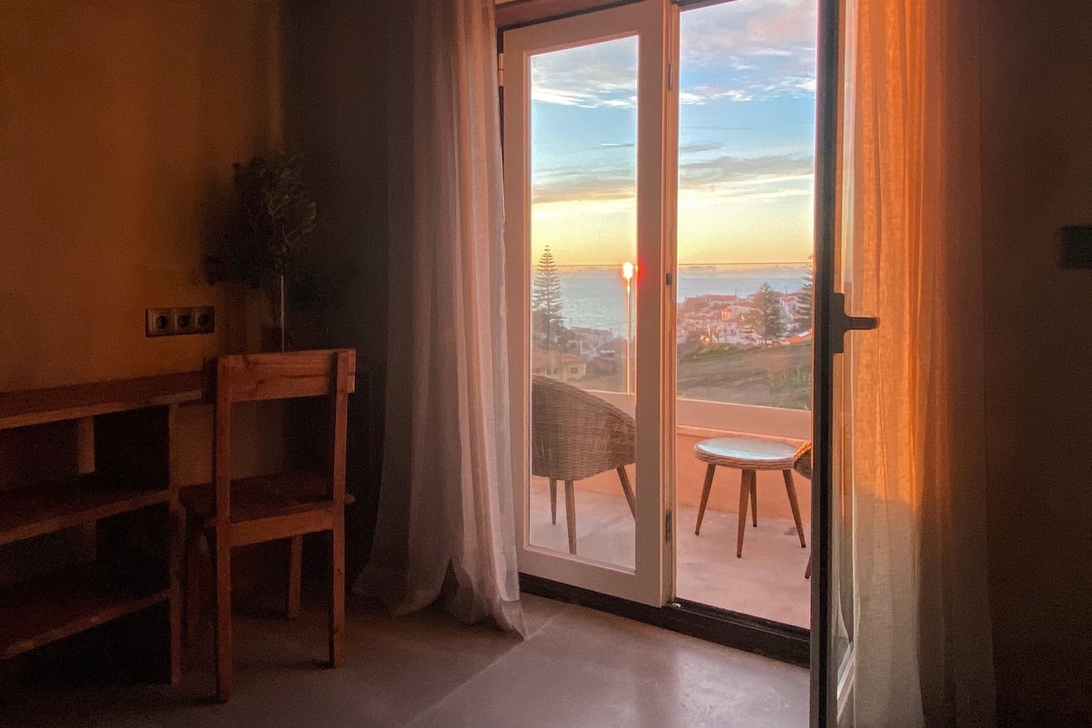 Quarto com um banco de madeira, uma cadeira e vista para o pôr do sol sobre a costa.