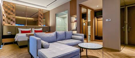 Quarto de hotel moderno com um sofá modular cinzento, uma mesa lateral redonda, um espelho grande e piso de madeira.