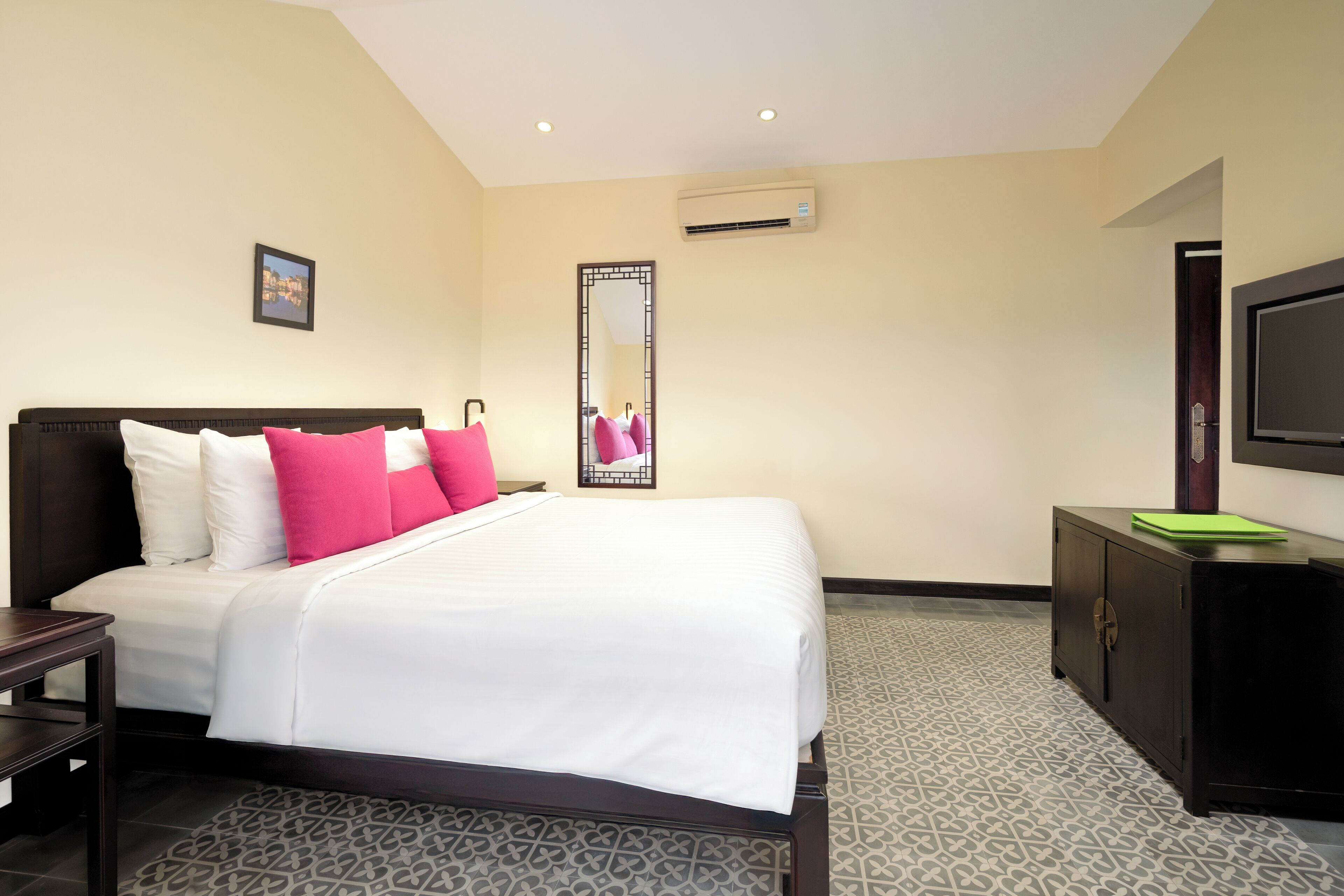 Quarto de hotel com uma cama grande, mesa de cabeceira, cômoda, espelho, aparelho de ar condicionado e televisão.