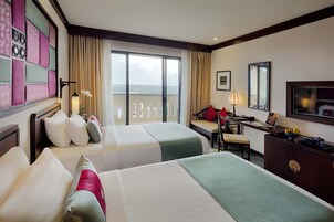 Quarto de hotel com duas camas, uma escrivaninha, uma cadeira, televisão e varanda com vista.
