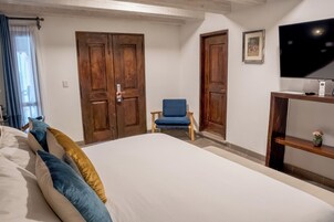 Quarto de hotel com uma cama grande, duas portas de madeira, uma cadeira azul, uma televisão e uma pequena prateleira com um vaso.
