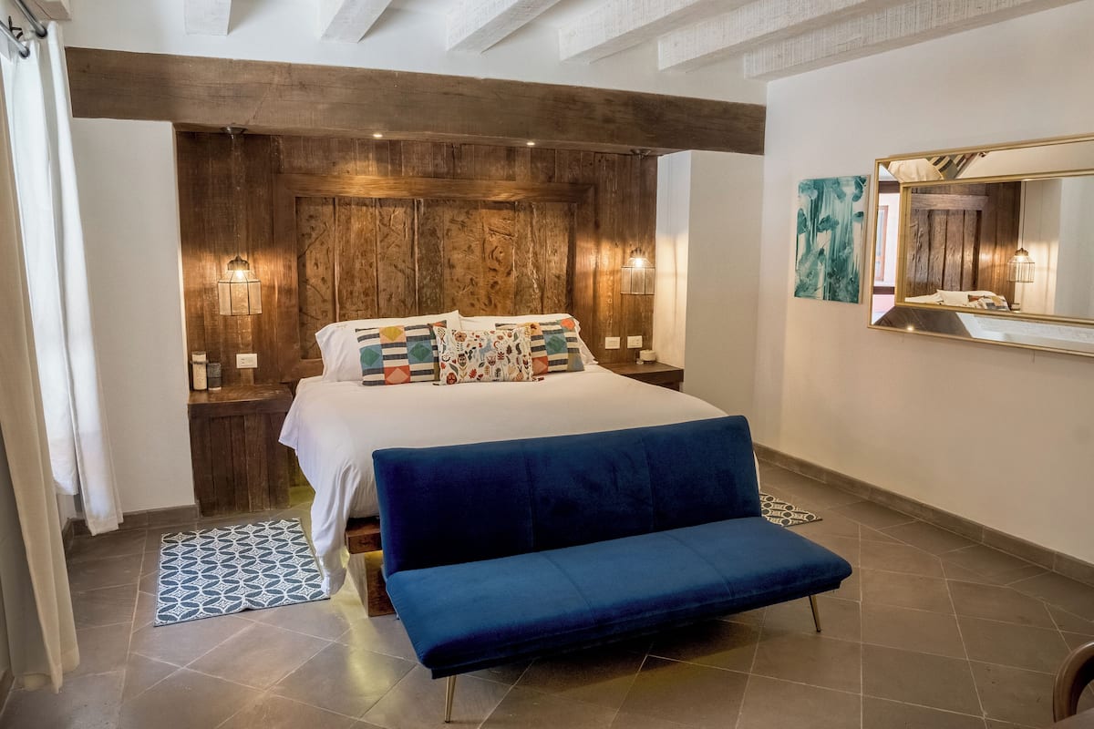 Um quarto com cama, mesas de cabeceira, um banco azul, tapete estampado, cabeceira de madeira e um espelho.