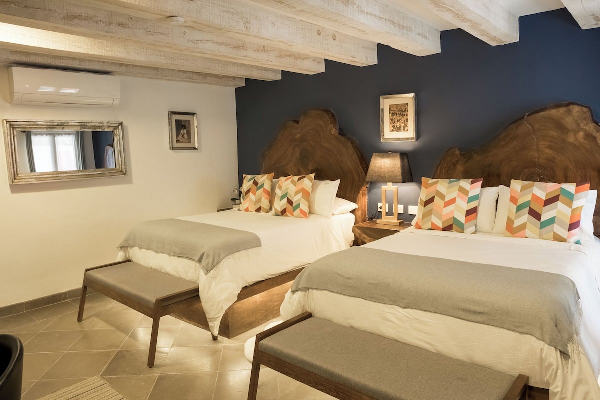 Quarto de hotel com duas camas, uma cabeceira de madeira, uma mesinha de cabeceira, uma luminária e um espelho.