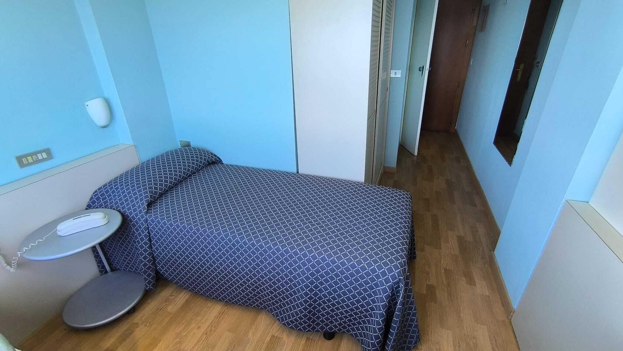 Quarto pequeno e compacto com uma cama de solteiro, uma mesinha redonda e uma luminária na parede.