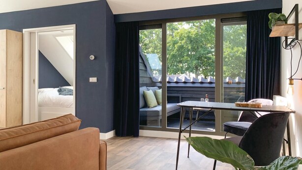 Quarto moderno com cama, sofá, mesa de jantar e uma grande janela com vista para árvores.