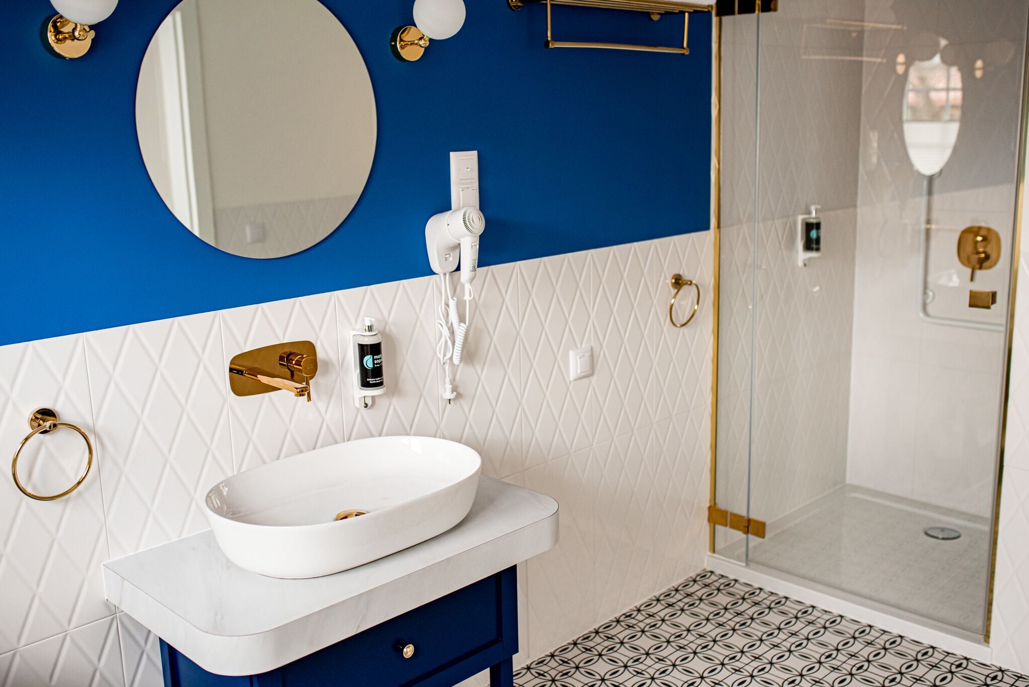Uma casa de banho com uma pia branca, azulejos azuis e brancos nas paredes e um cabine de chuveiro em vidro, com detalhes dourados.