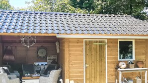 Uma cabana de madeira com telhado de telhas, uma porta e um pequeno alpendre com móveis de vime e uma mesa.