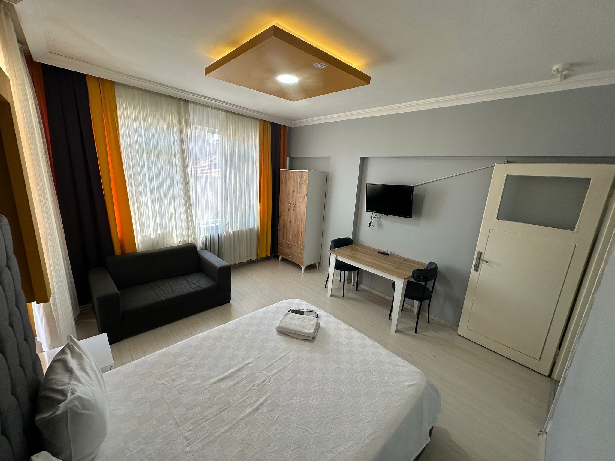 Quarto de hotel com cama, escrivaninha, sofá, guarda-roupa e televisão fixada na parede.