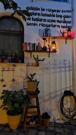 Uma área de estar ao ar livre aconchegante, com plantas em vasos, um banquinho de madeira e uma prateleira com livros e um candeeiro.