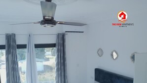 Um quarto com ventilador de teto, janelas amplas com cortinas, uma cama com cabeceira escura, e o logotipo de The View Apartments Gambia na parede.