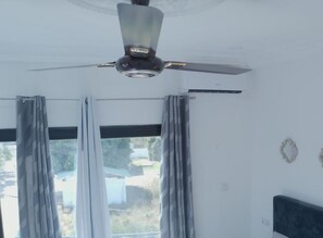 Um quarto com ventilador de teto, janelas amplas com cortinas, uma cama com cabeceira escura, e o logotipo de The View Apartments Gambia na parede.
