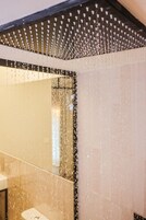 Chuveiro com recurso de água tipo chuva, um espelho e parede revestida com azulejos bege.