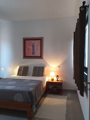Quarto com cama, mesa de cabeceira com abajur, quadro na parede e janela com cortinas.