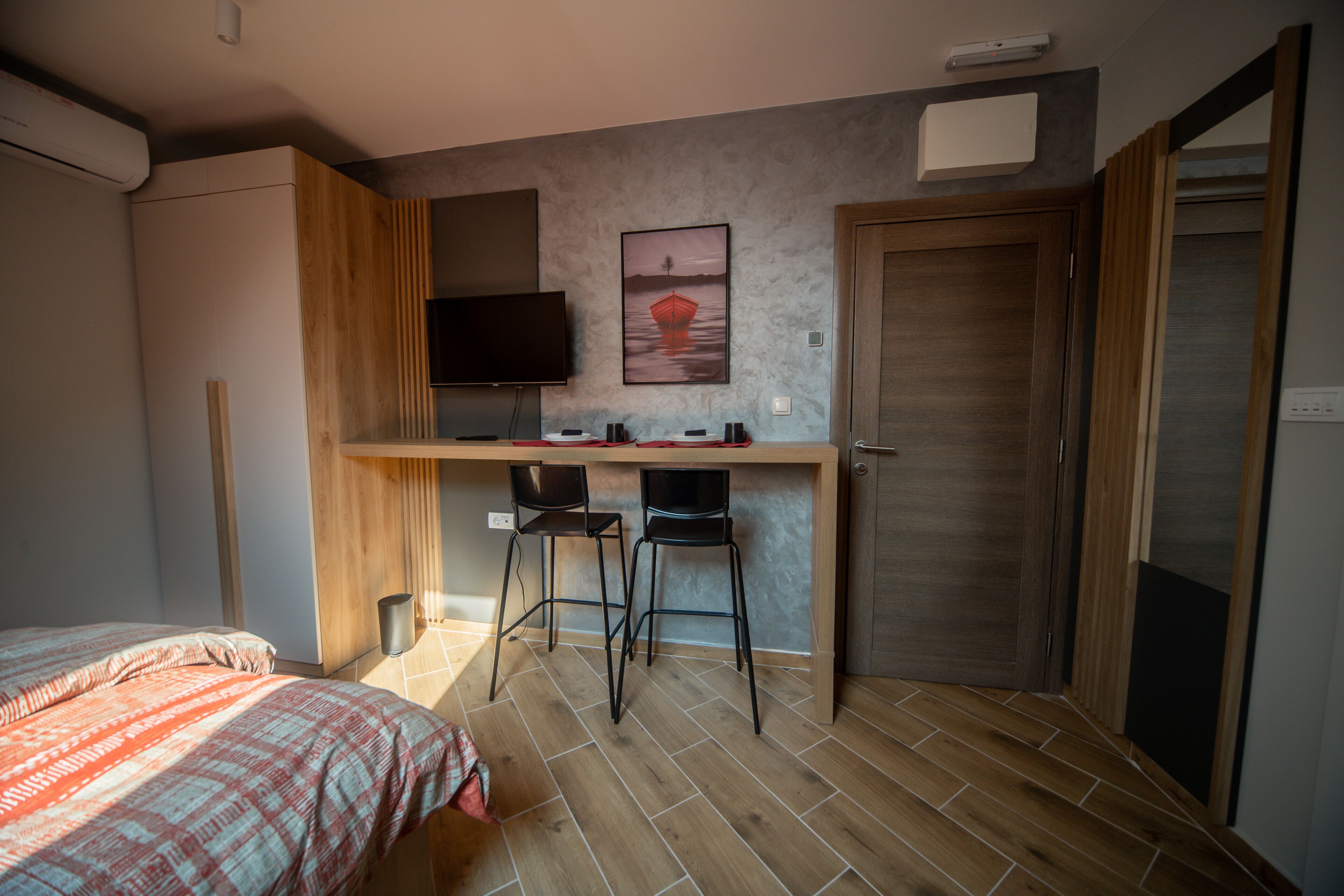 Quarto de hotel compacto com uma cama, uma escrivaninha com duas cadeiras, uma TV de tela plana e uma porta.